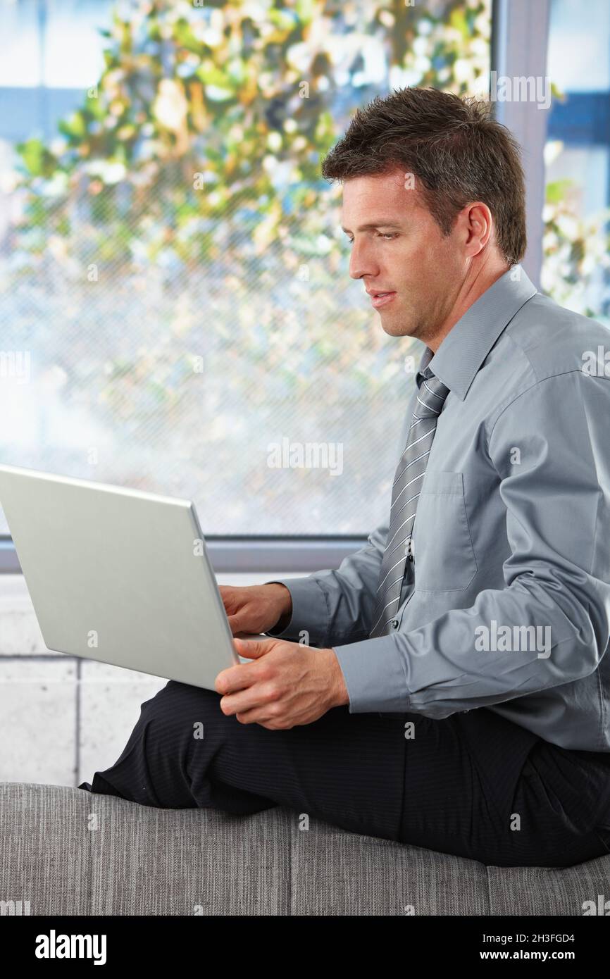 Businessman on couch using his laptop smiling at camera in the office ...