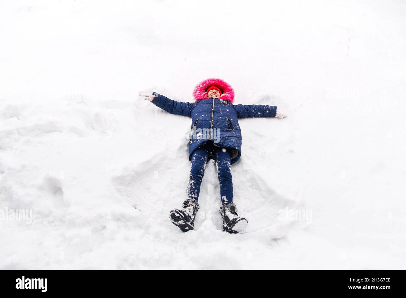 Preteen smiling girl in bright pink warm hood making snow angel while ...