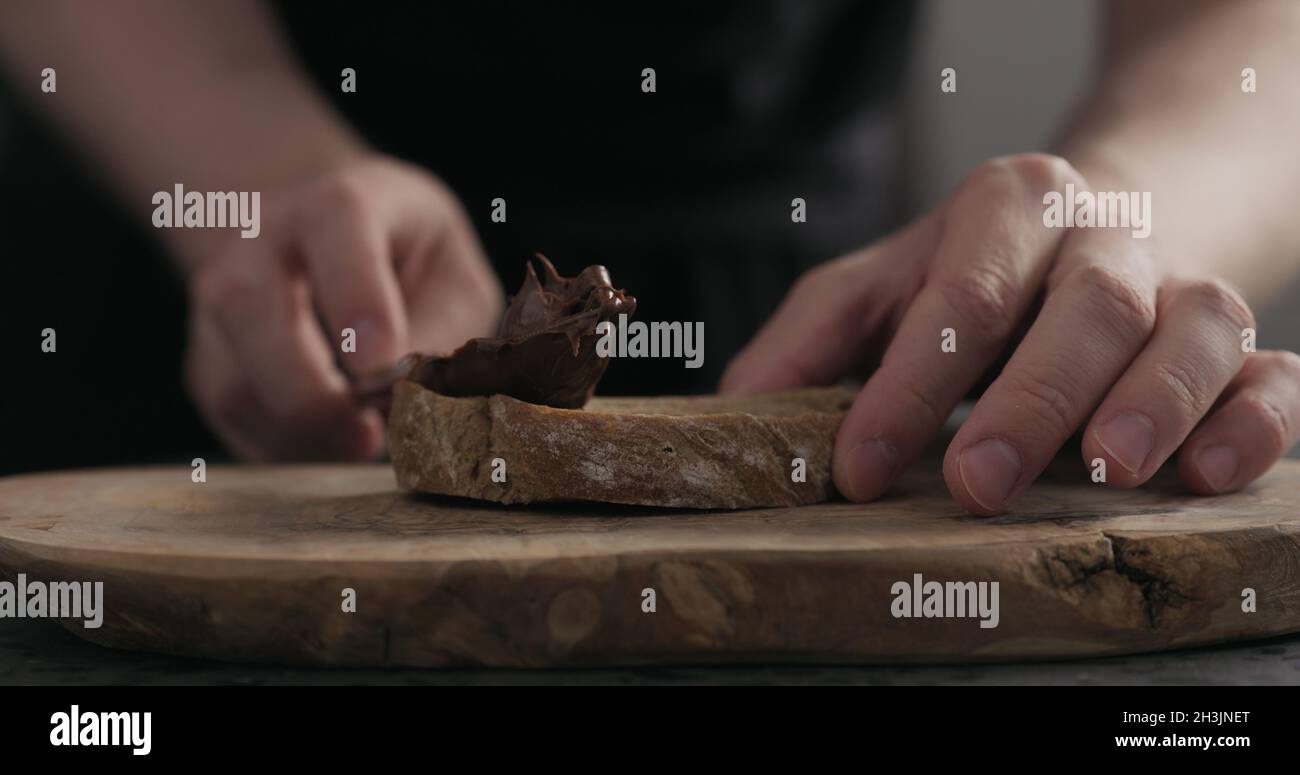 man spreading chocolate hazelnut spread on ciabatta slice on wood board ...