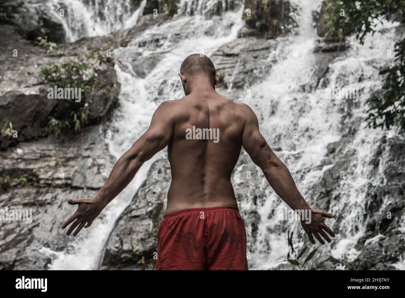 Back view of muscular man standing with raised arms near waterfall ...