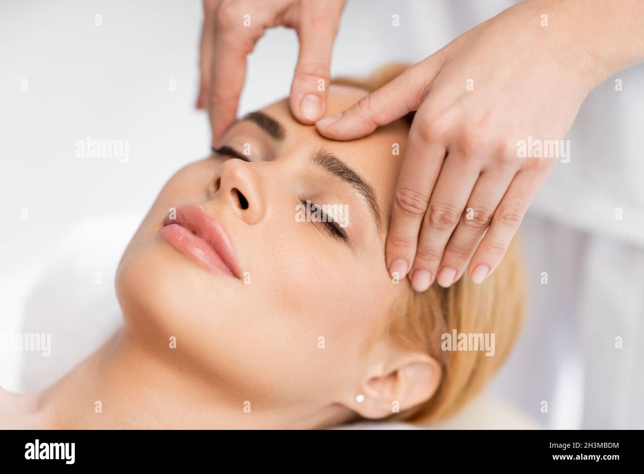 close up of blonde client with closed eyes receiving face massage in ...
