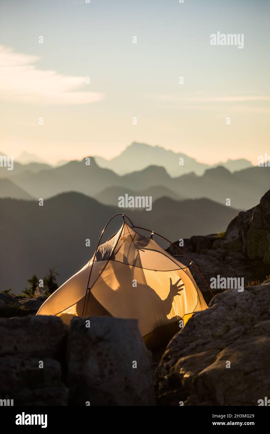 Man in tent reaches hand out, shadow of fingers toward mountain peaks ...