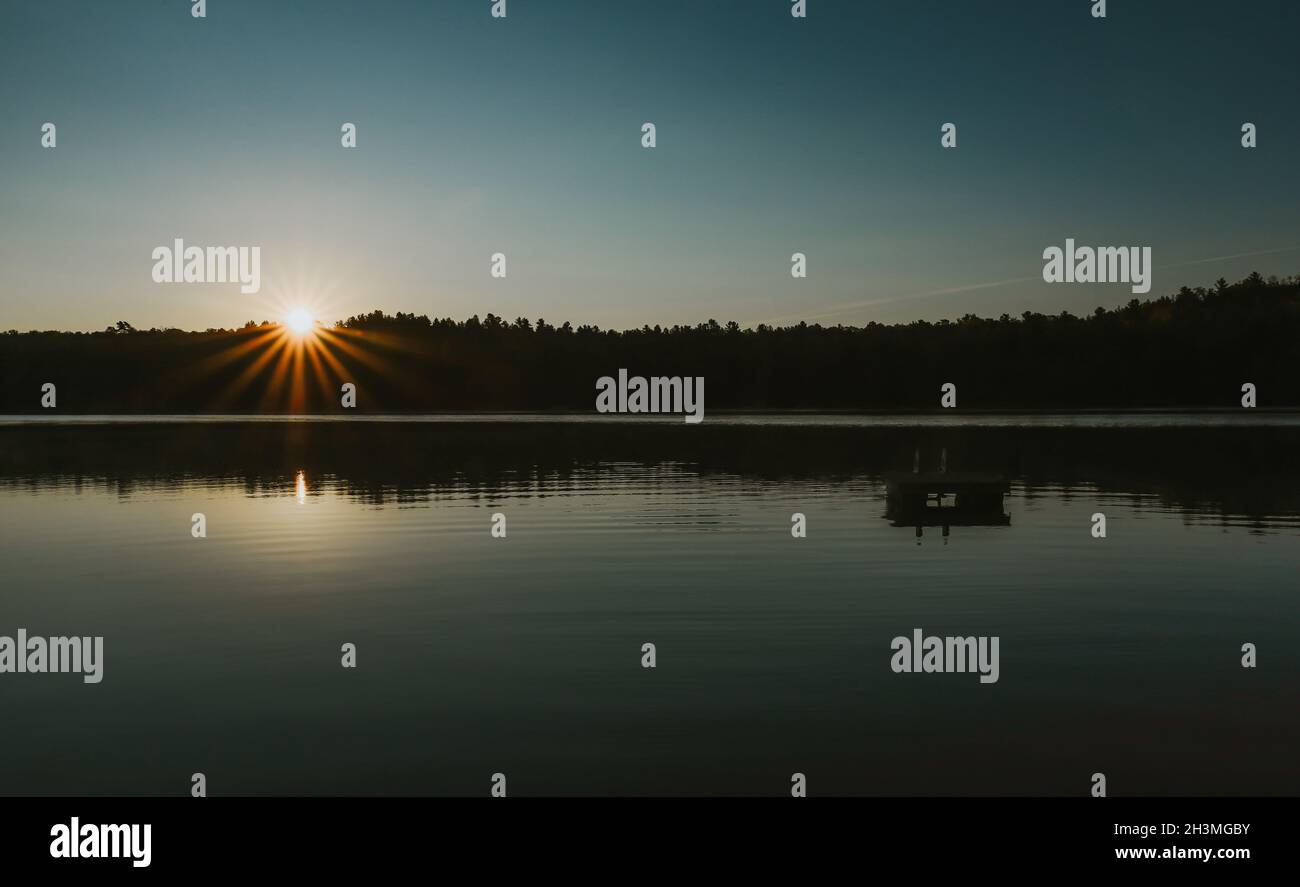 Sun rising on the tree lined horizon of a still lake Stock Photo - Alamy