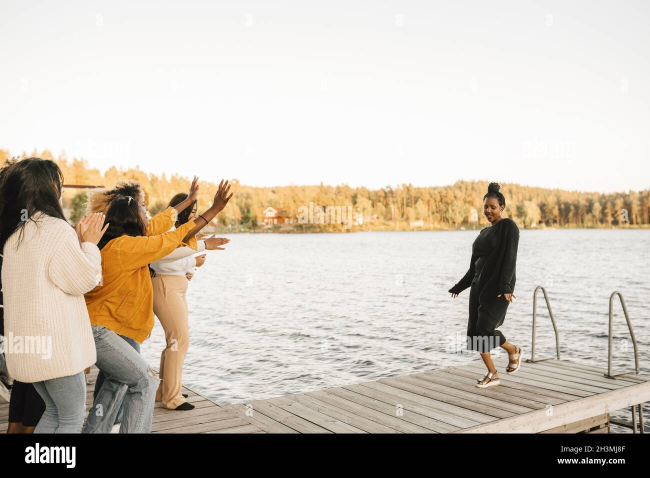 Woman dancing on jetty while female friends cheering at lakeshore ...