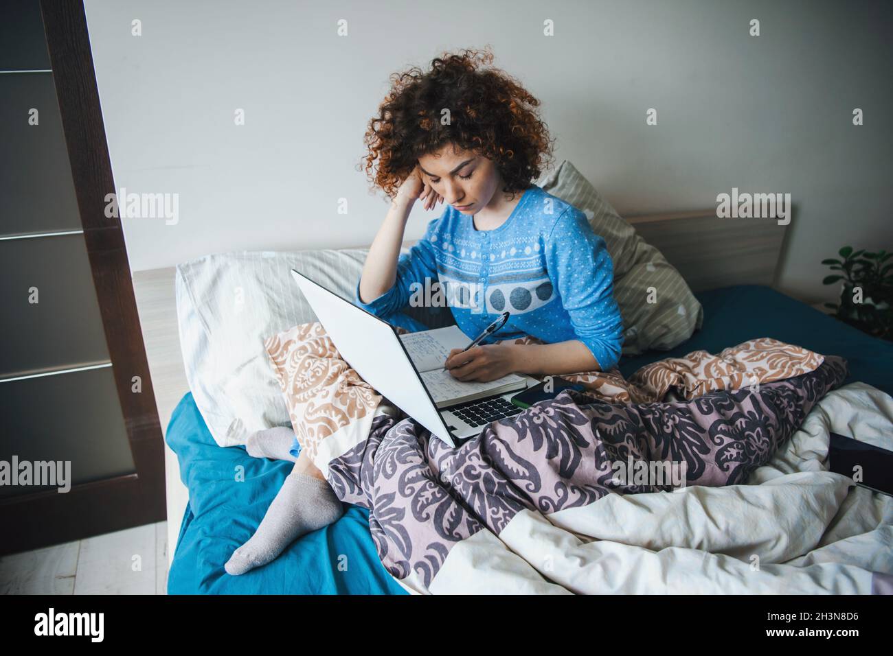 Curly woman studying remotely sitting in untidy bed. Modern woman ...