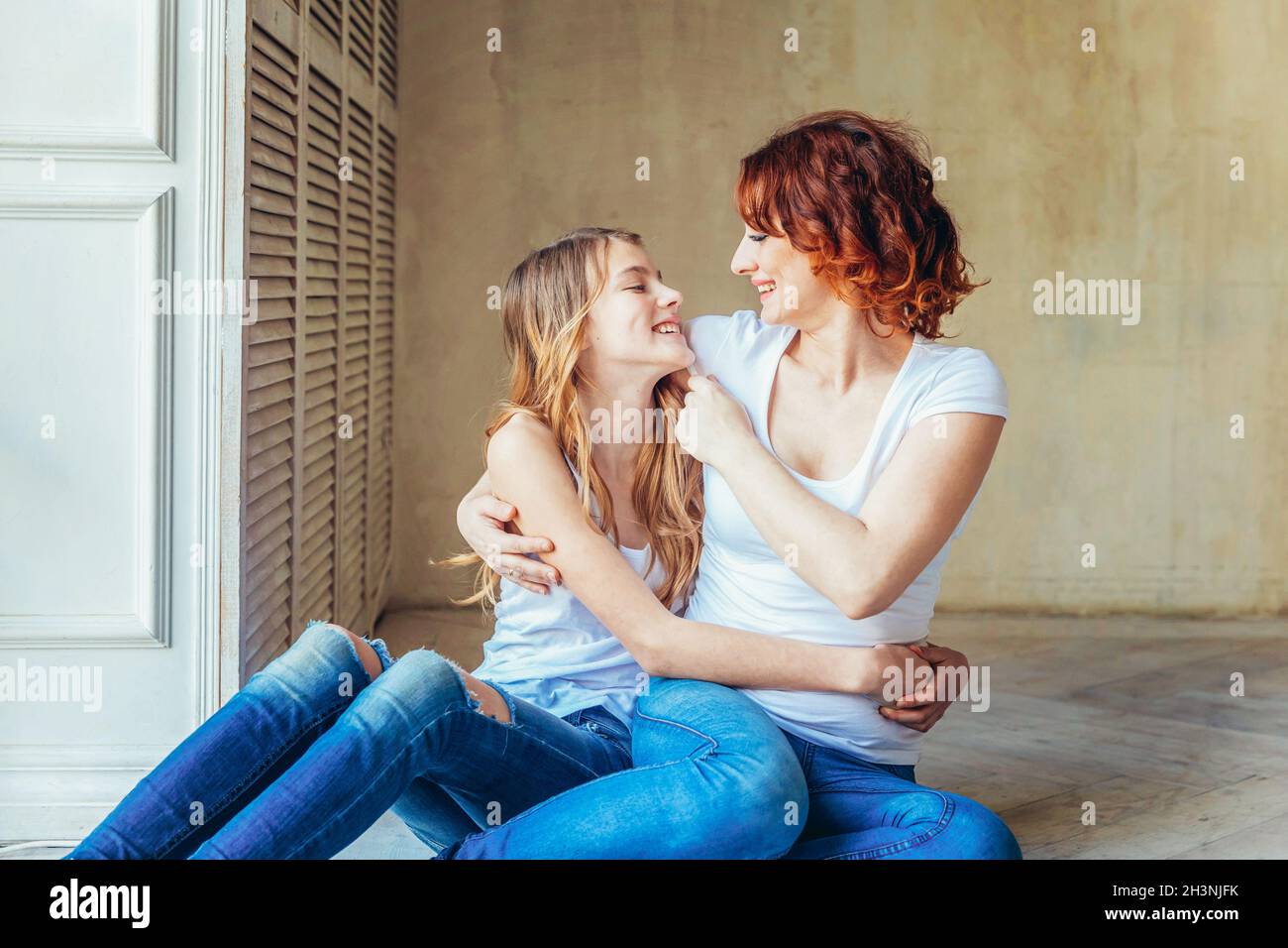 Young mother embracing her child. Woman and teenage girl relaxing in white bedroom near gray ...