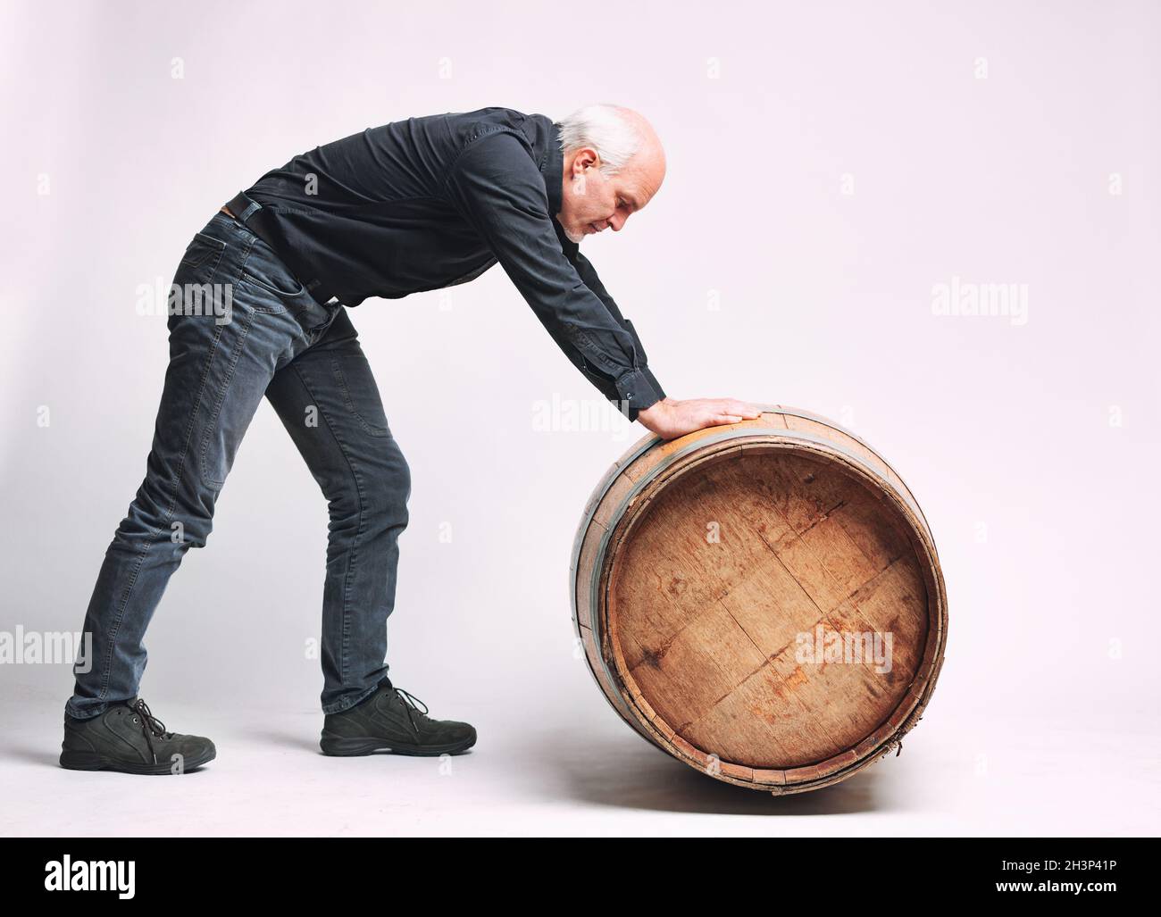 Senior man rolling an old oak wine or beer barrel in front of him along ...