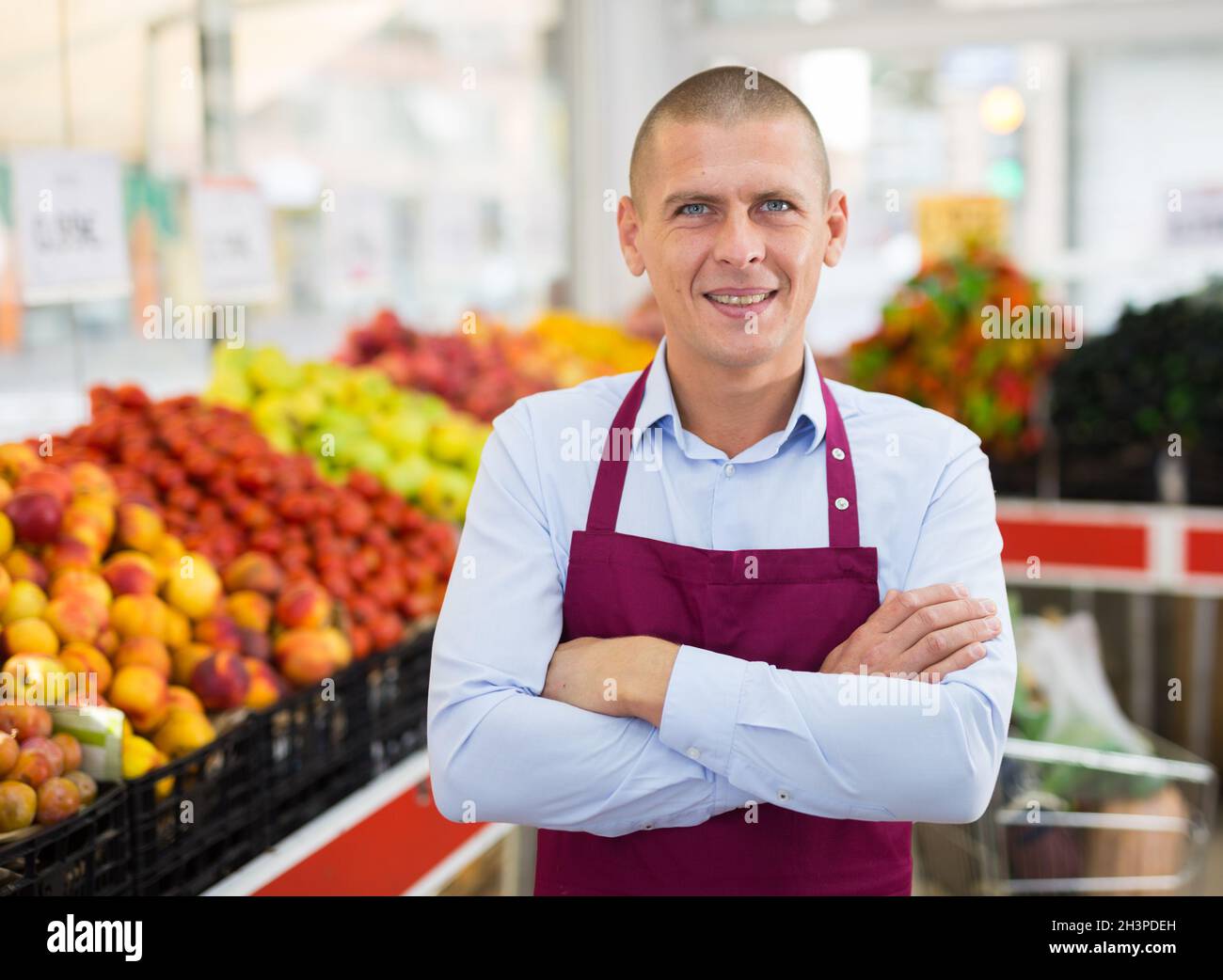 Confident greengrocery owner near shelves with fruits and vegetables ...