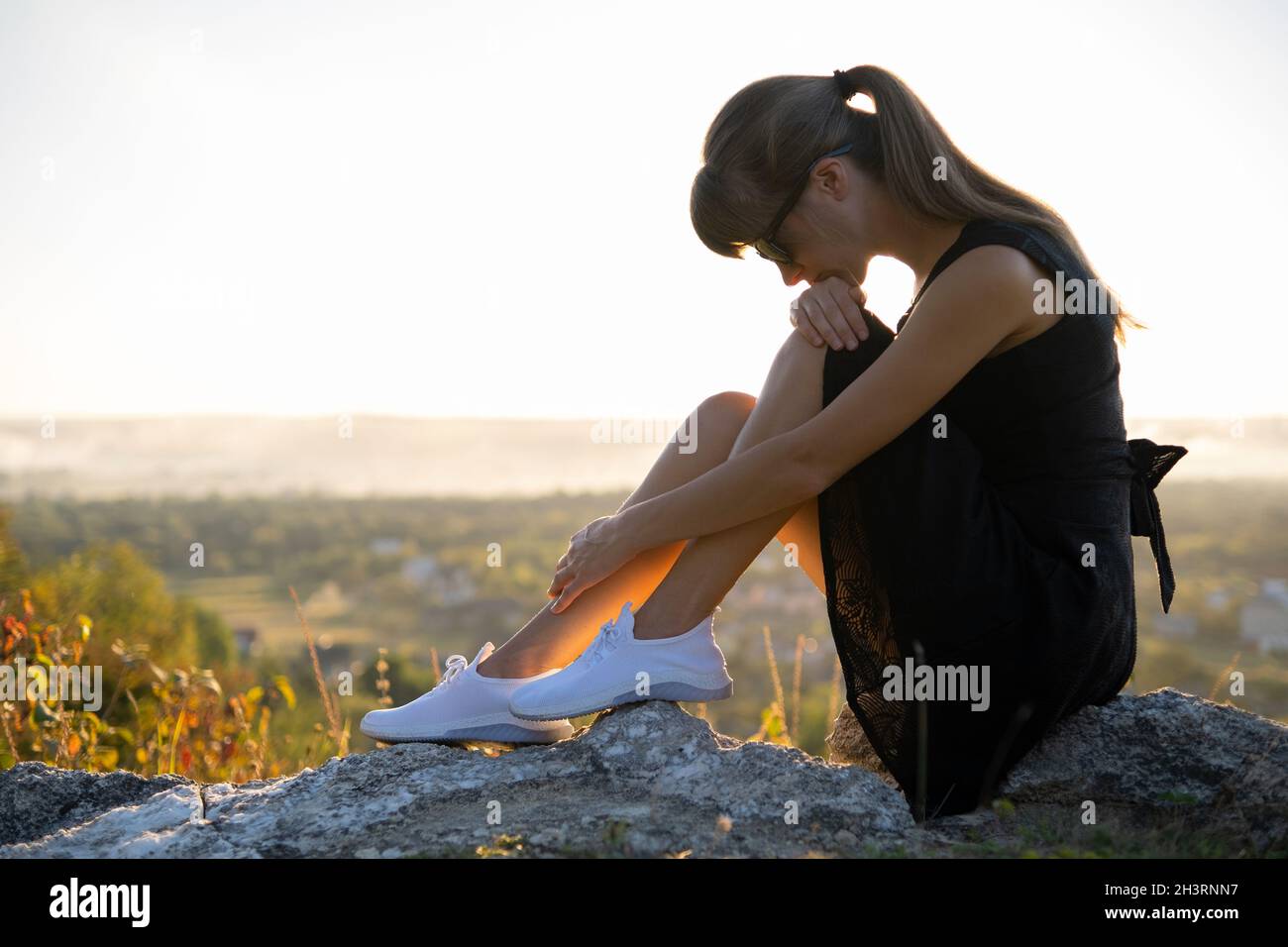 Pretty sad woman in black short summer dress sitting on a rock thinking ...