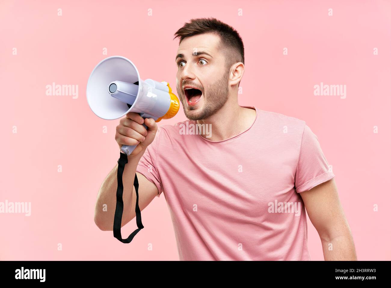 Funny young man shouting loud holding a megaphone over pink background ...