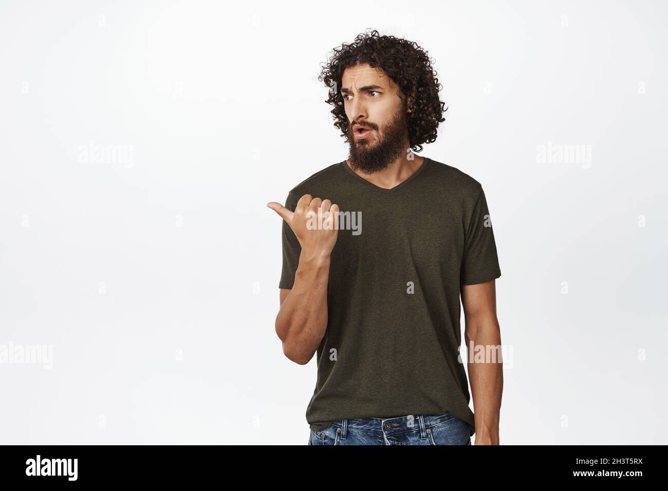 Confused young bearded turkish guy, pointing and looking left with ...