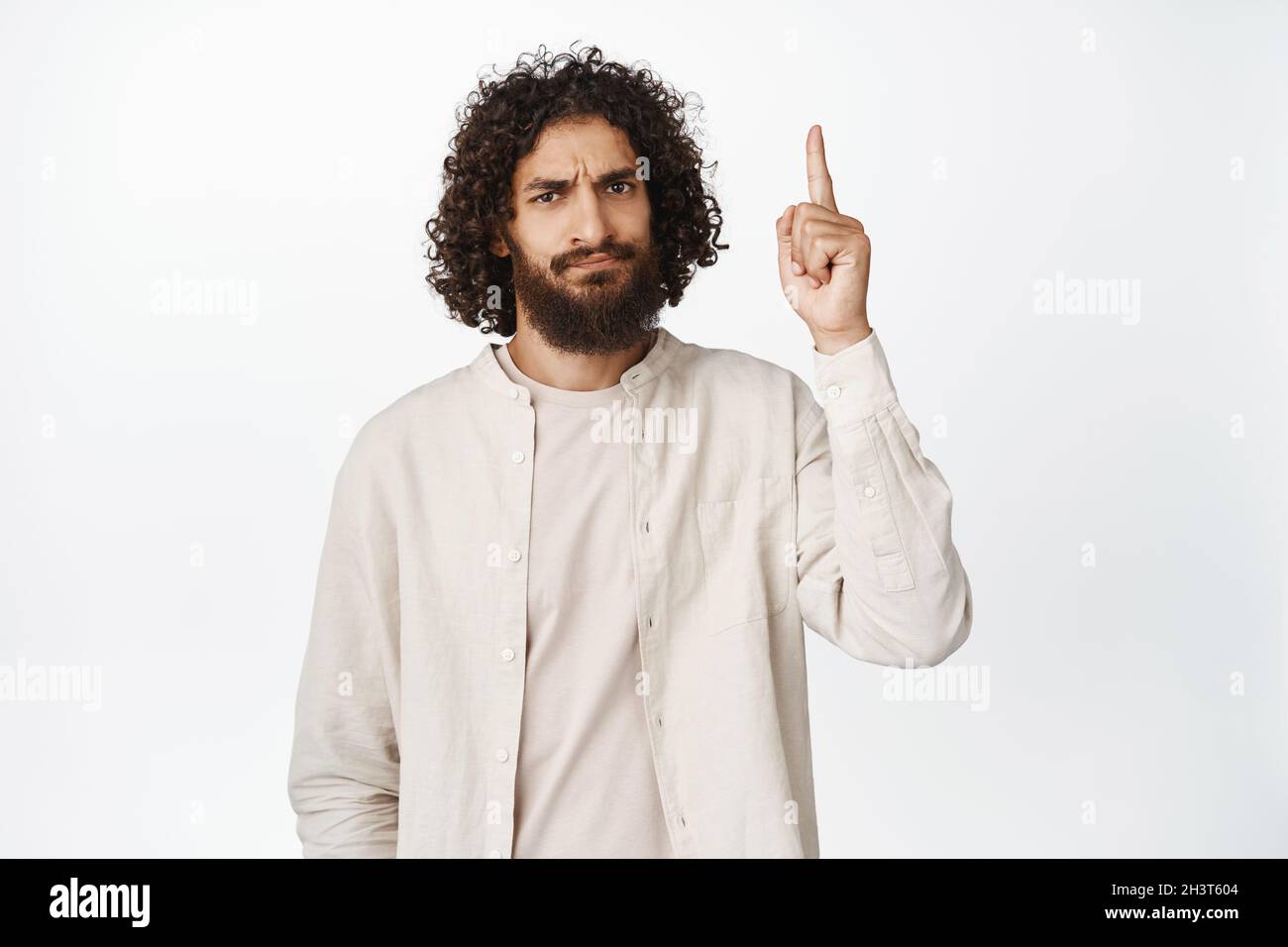 Suspicious young man pointing finger up, looking doubtful at camera ...