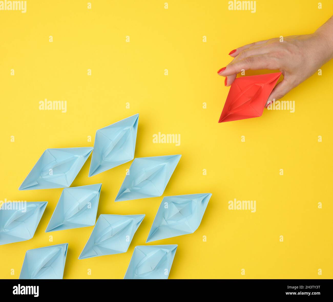A group of blue paper boats follows a red boat in front of a yellow ...