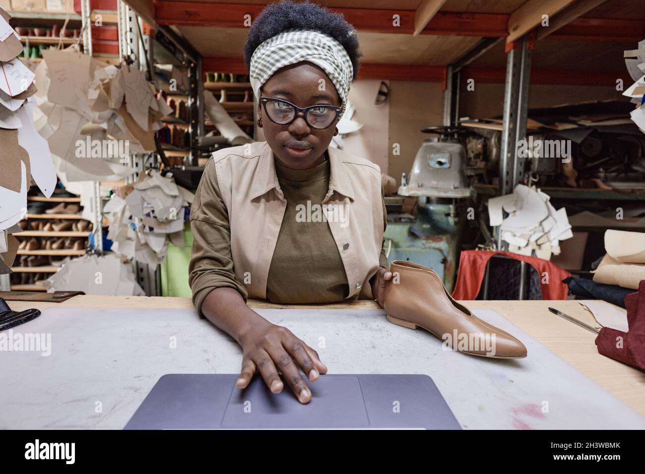 Serious African shoemaker sitting using laptop at her workplace while ...