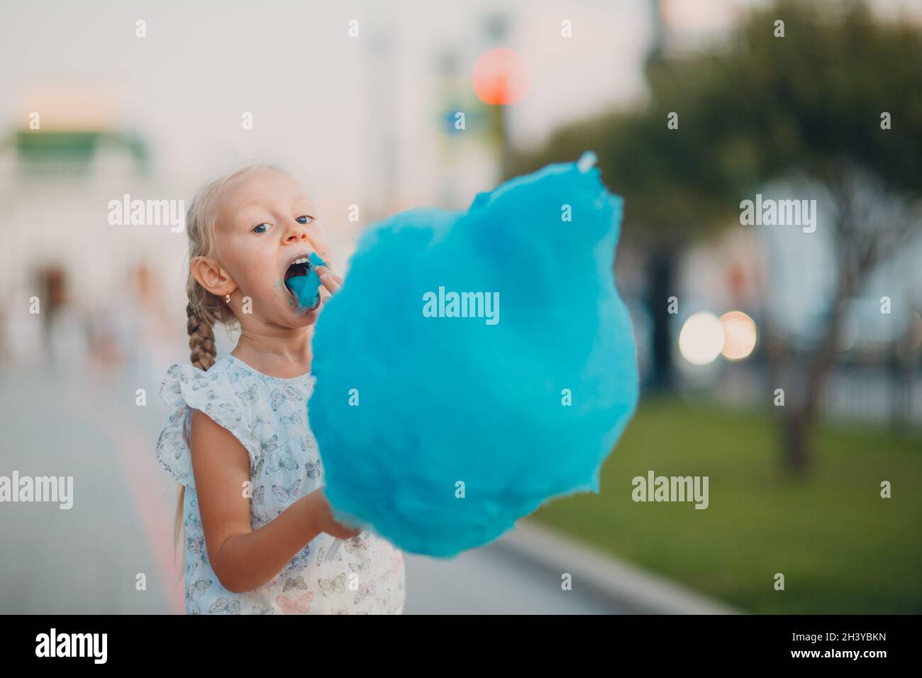 Little blond girl eating cotton candy and shows blue tongue in the park ...