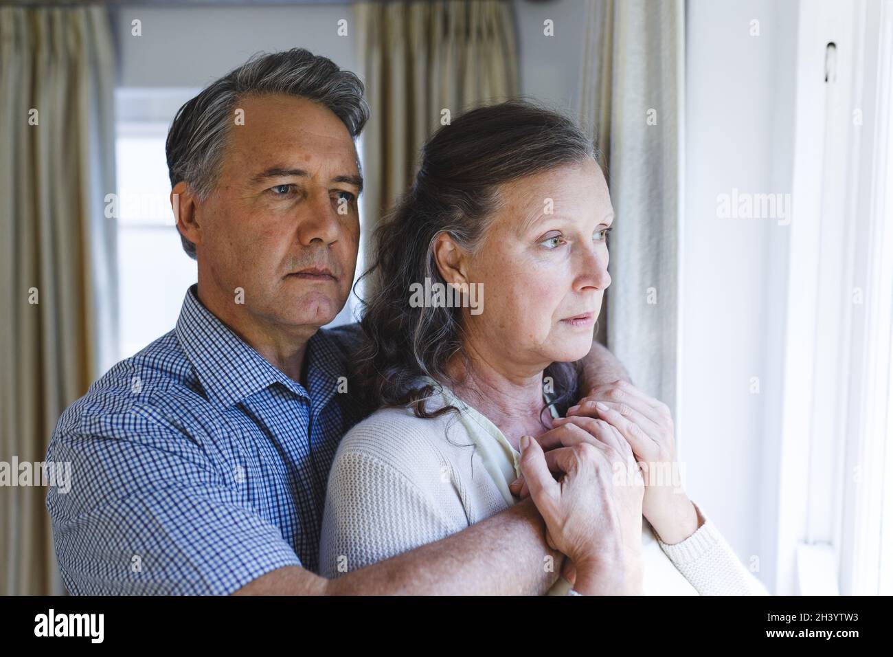 Thoughtful senior caucasian couple standing next to window, embracing ...