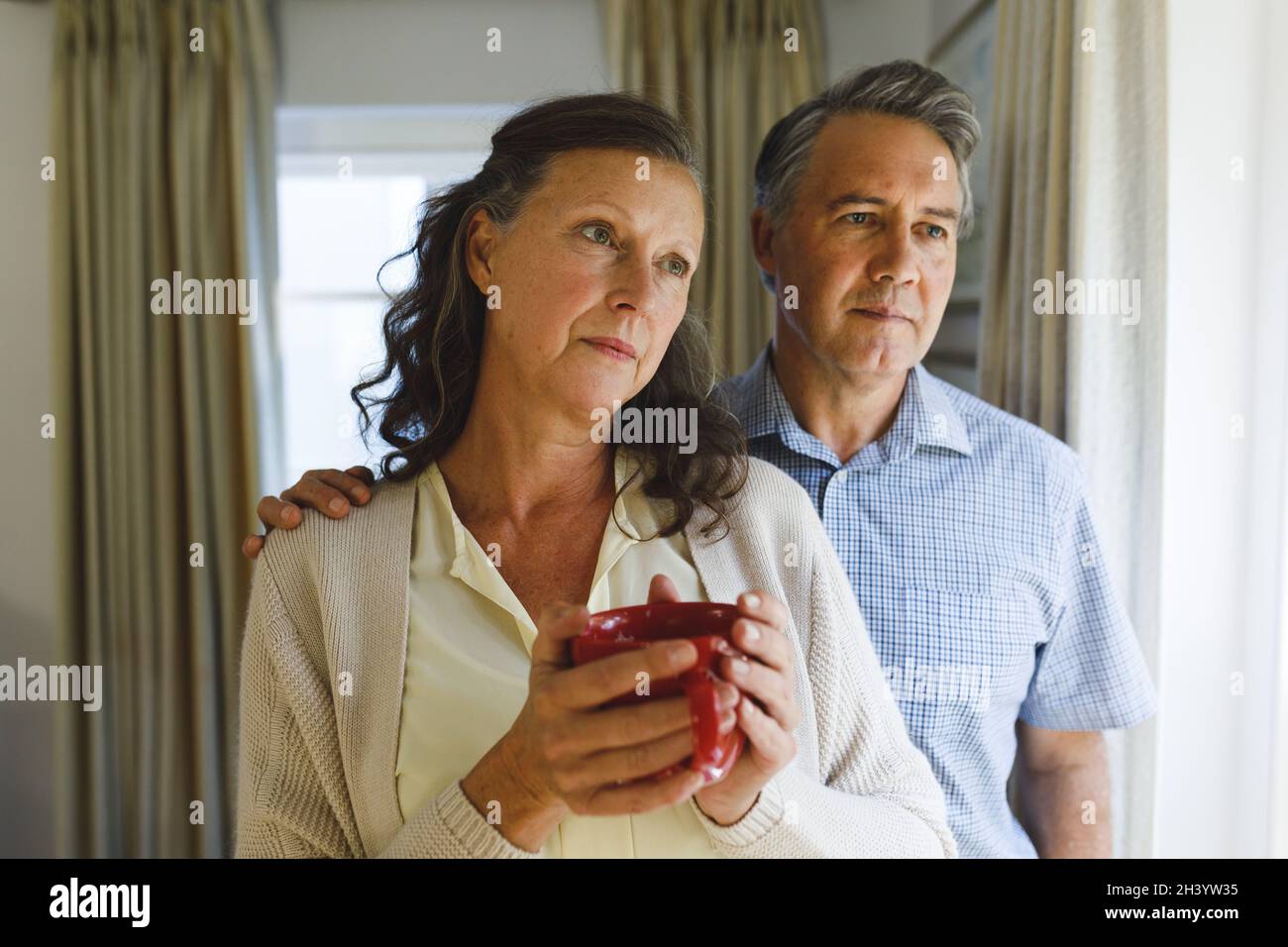 Happy senior caucasian couple standing next to window, holding cups ...