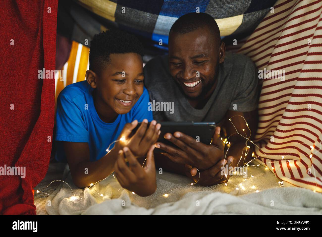 Happy african american father and son lying in blanket fort, using ...