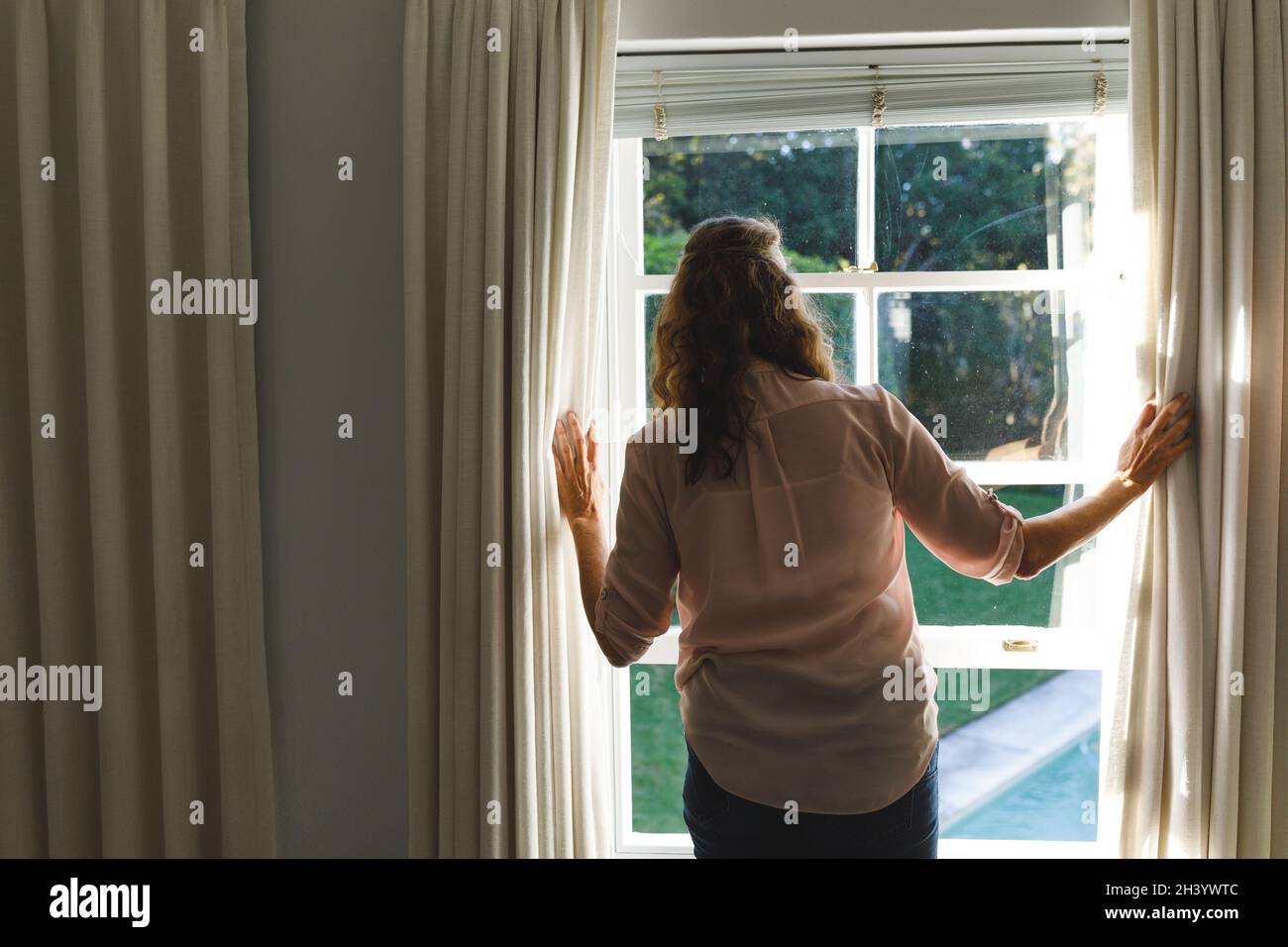 Thoughtful senior caucasian woman in bedroom, standing next to window ...