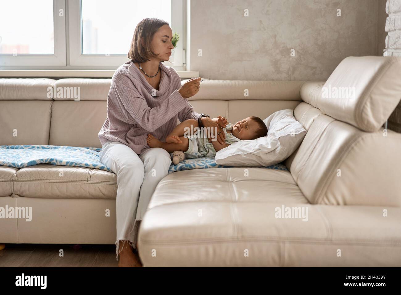 Worried young mother sitting on sofa beside her sick son with high fever. Mom measures ...