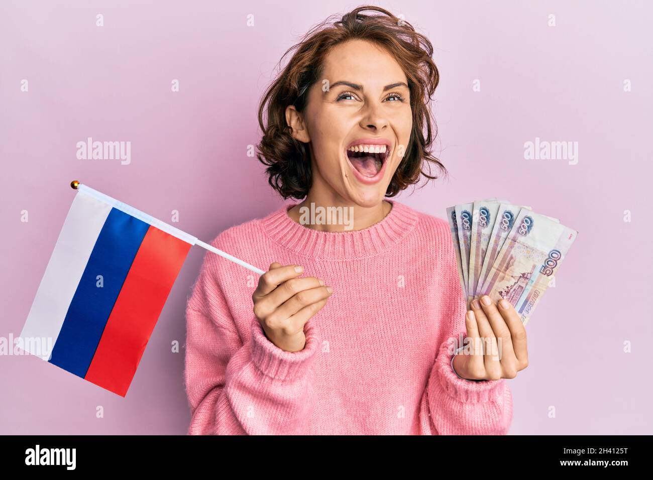 Young brunette woman holding russia flag and rubles banknotes angry and ...