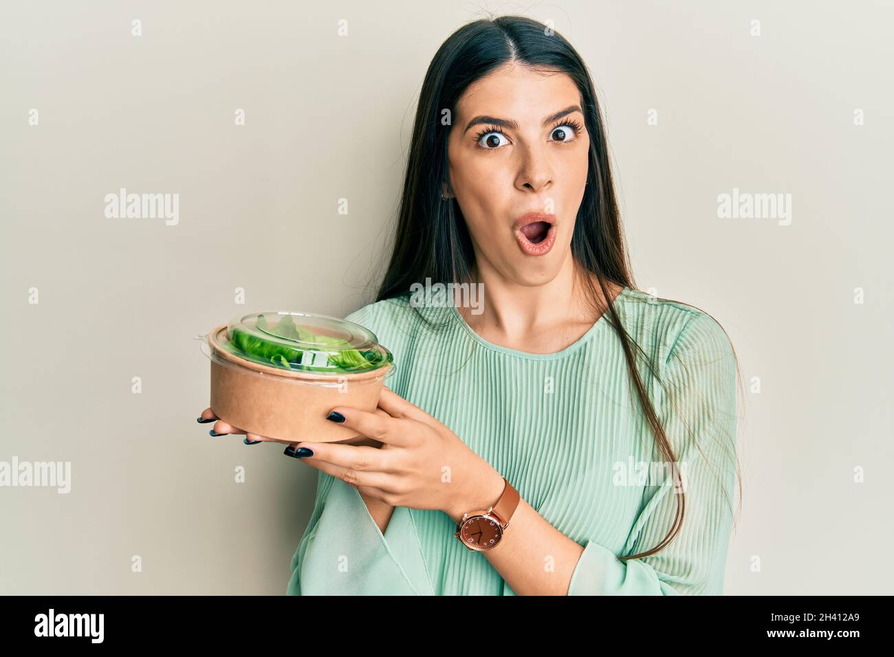 Young hispanic woman eating take away salad scared and amazed with open ...