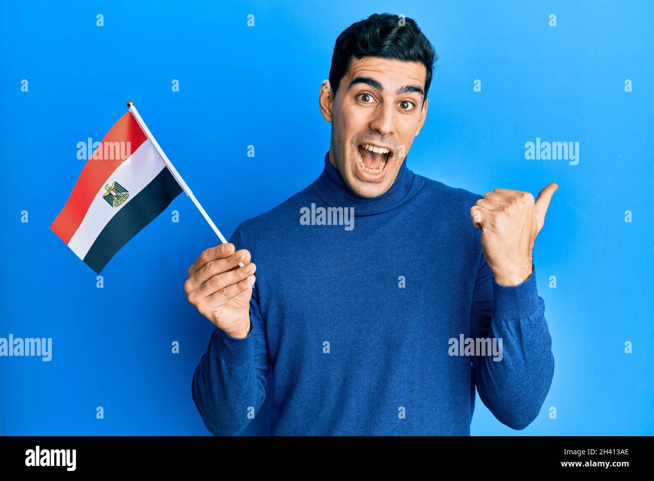 Handsome hispanic man holding egypt flag pointing thumb up to the side ...
