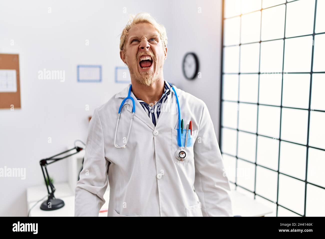 Young blond man wearing doctor uniform and stethoscope at clinic angry ...