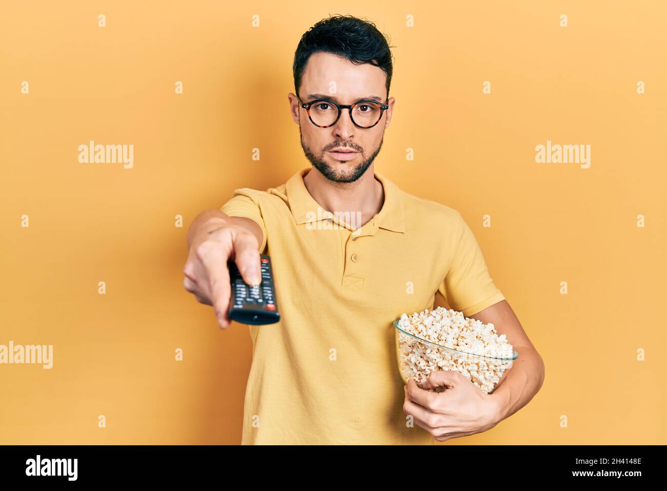 Young hispanic man eating popcorn using tv control skeptic and nervous ...