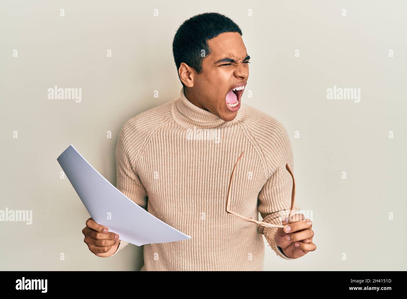 Young handsome hispanic man holding blank paper and glasses angry and ...