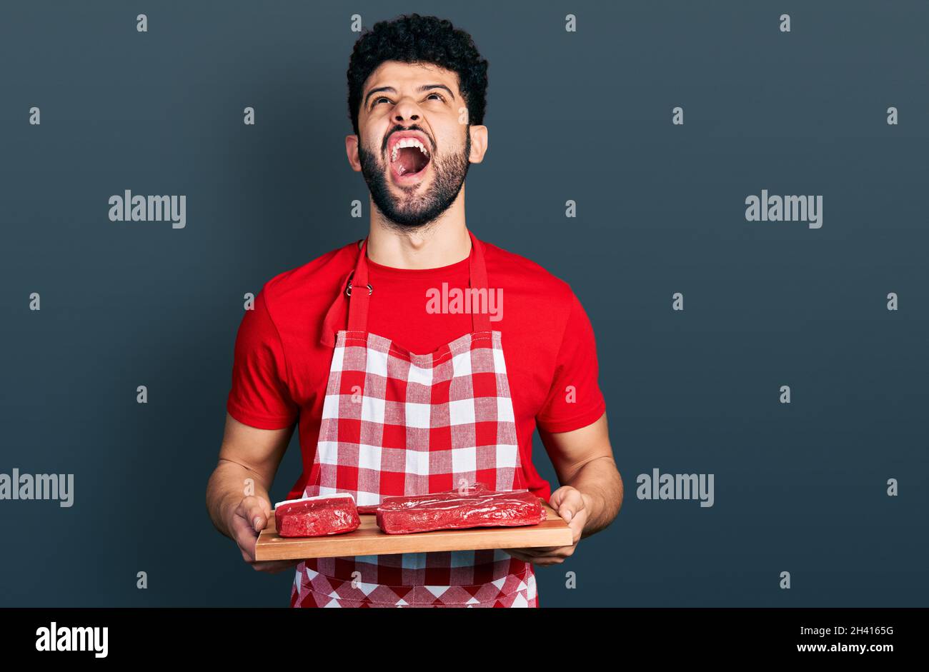 Young arab man with beard holding board with raw meat angry and mad ...