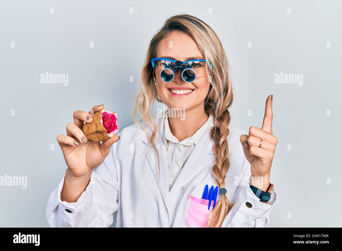 Beautiful young blonde woman holding geode stone wearing magnifier ...