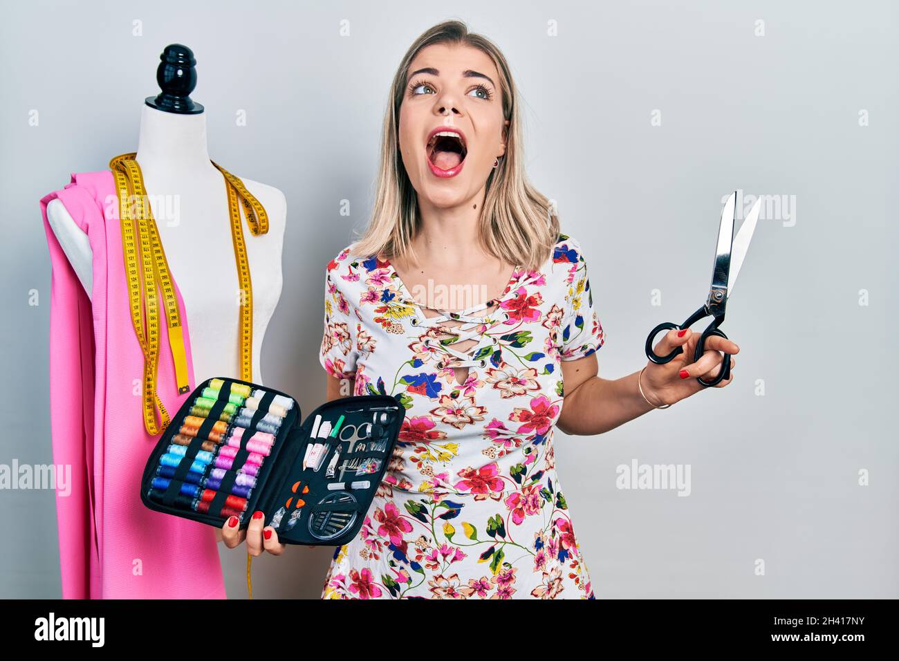 Beautiful caucasian woman dressmaker designer holding scissors and sewing kit angry and mad ...