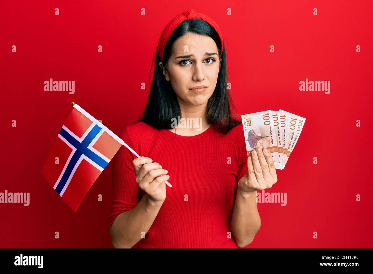 Young hispanic woman holding norway flag and krone banknotes depressed ...