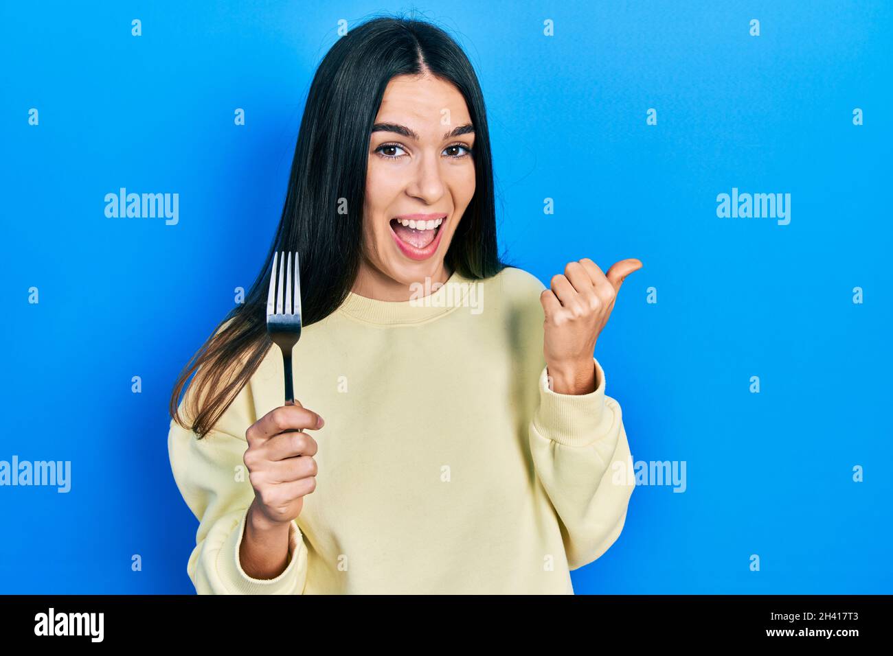 Young brunette woman holding one silver fork pointing thumb up to the ...