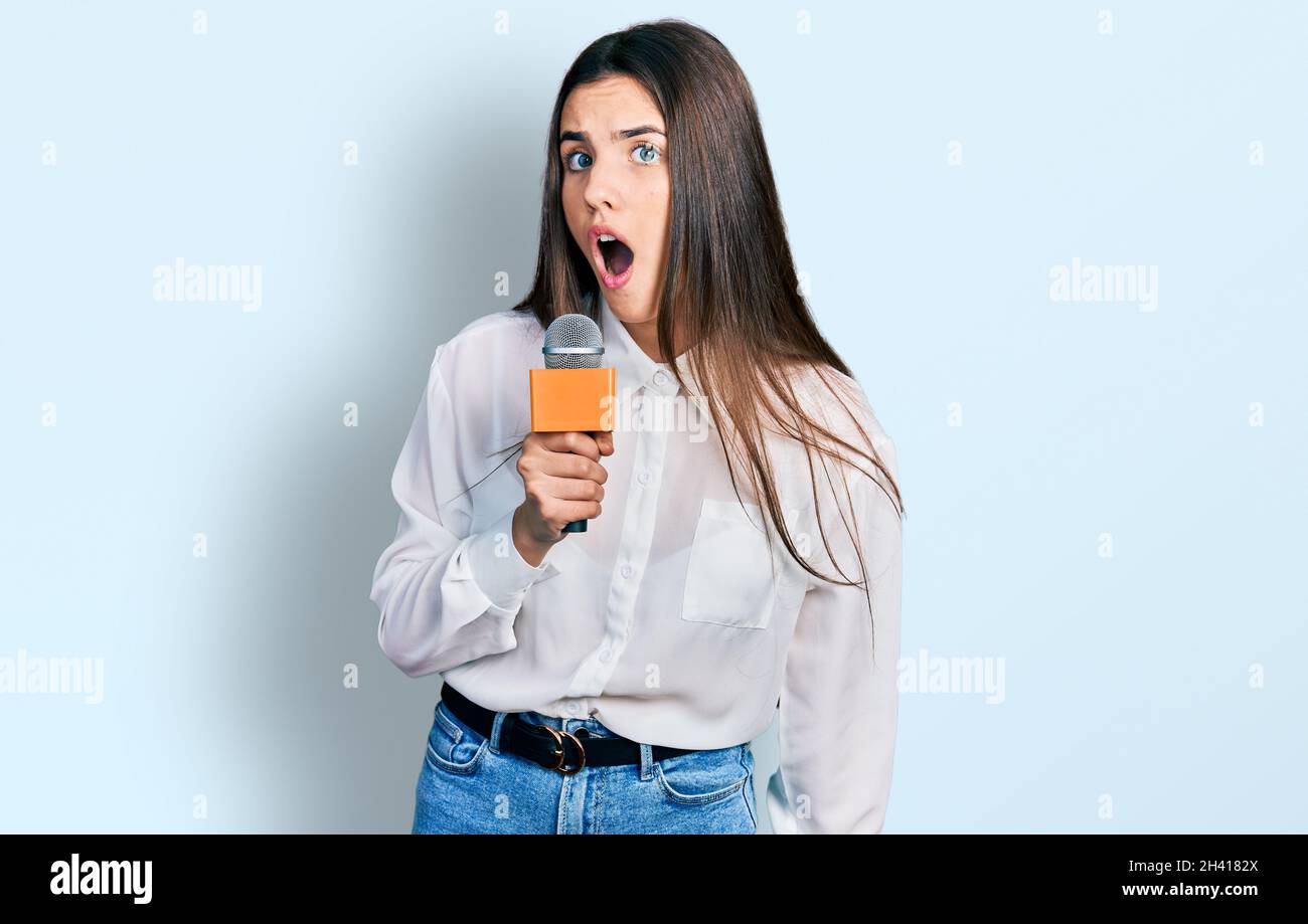 Young brunette teenager holding reporter microphone scared and amazed ...