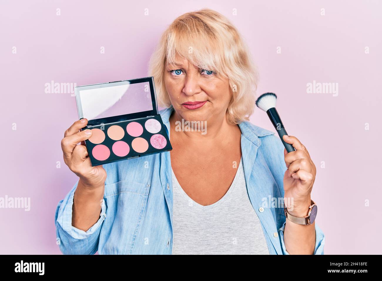 Middle age blonde woman holding makeup brush and blush skeptic and ...