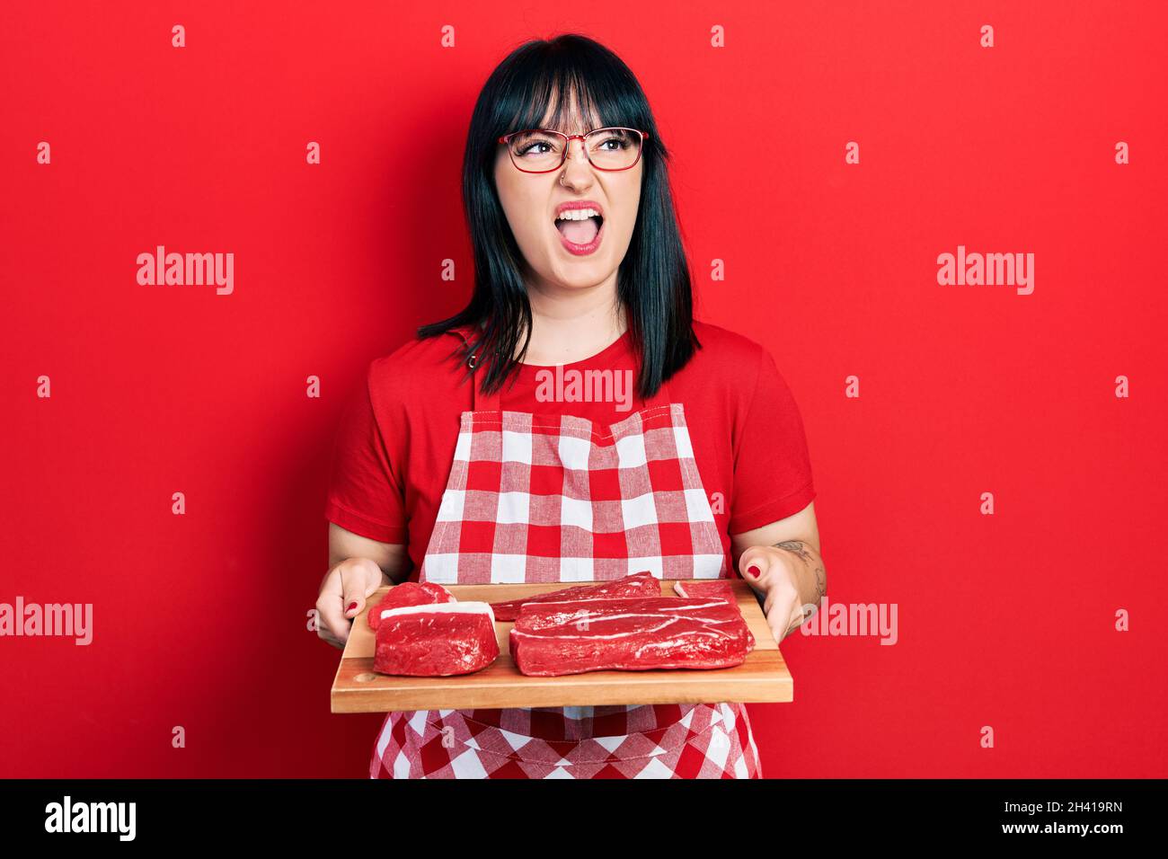 Young hispanic woman wearing cook apron holding board with meat angry and mad screaming ...