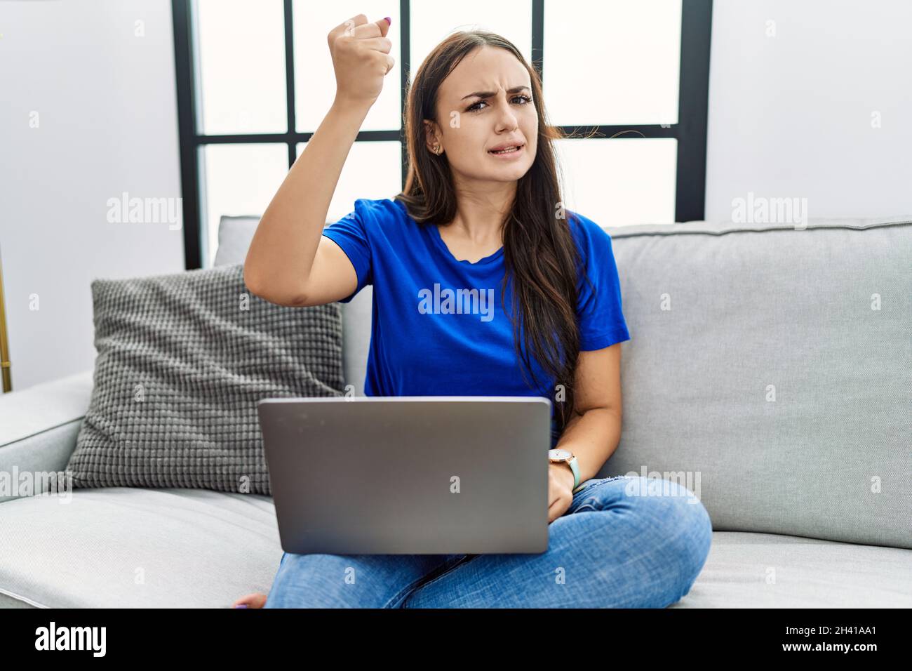 Young brunette woman using laptop at home angry and mad raising fist frustrated and furious ...