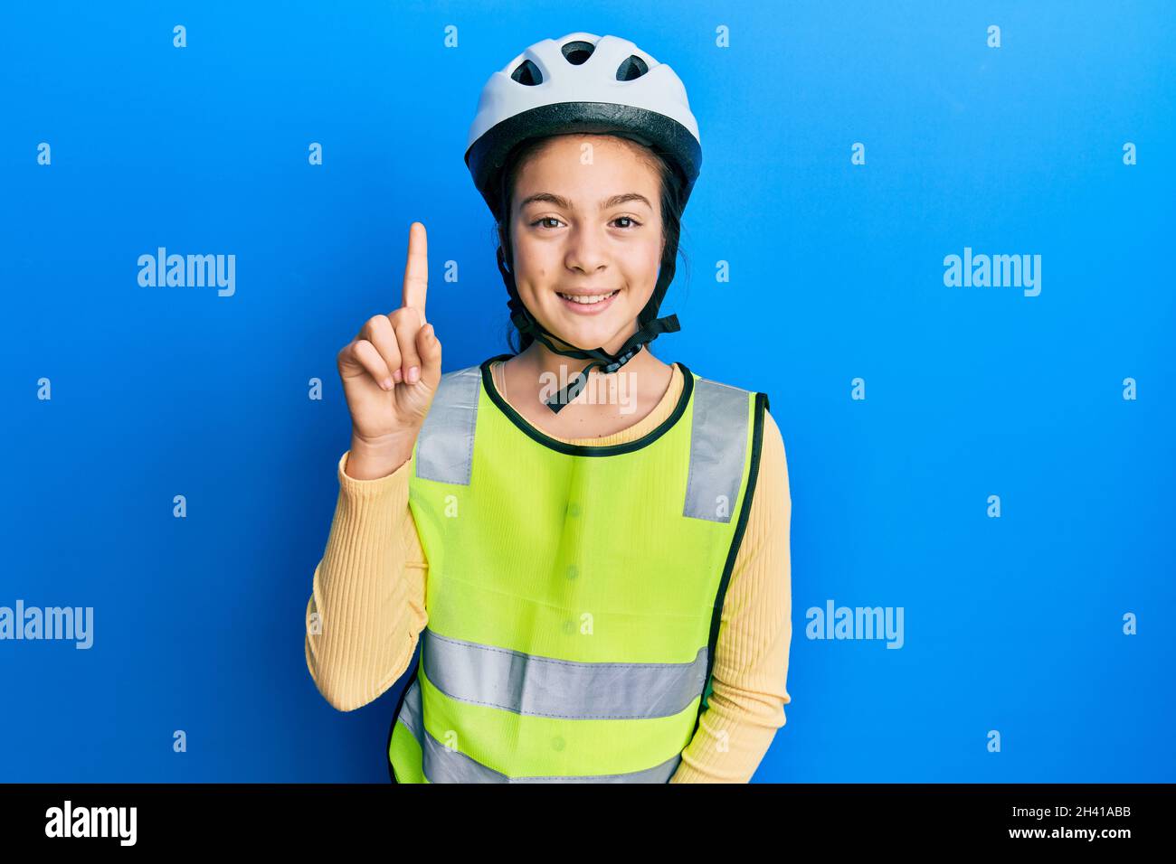 Beautiful brunette little girl wearing bike helmet and reflective vest showing and pointing up ...