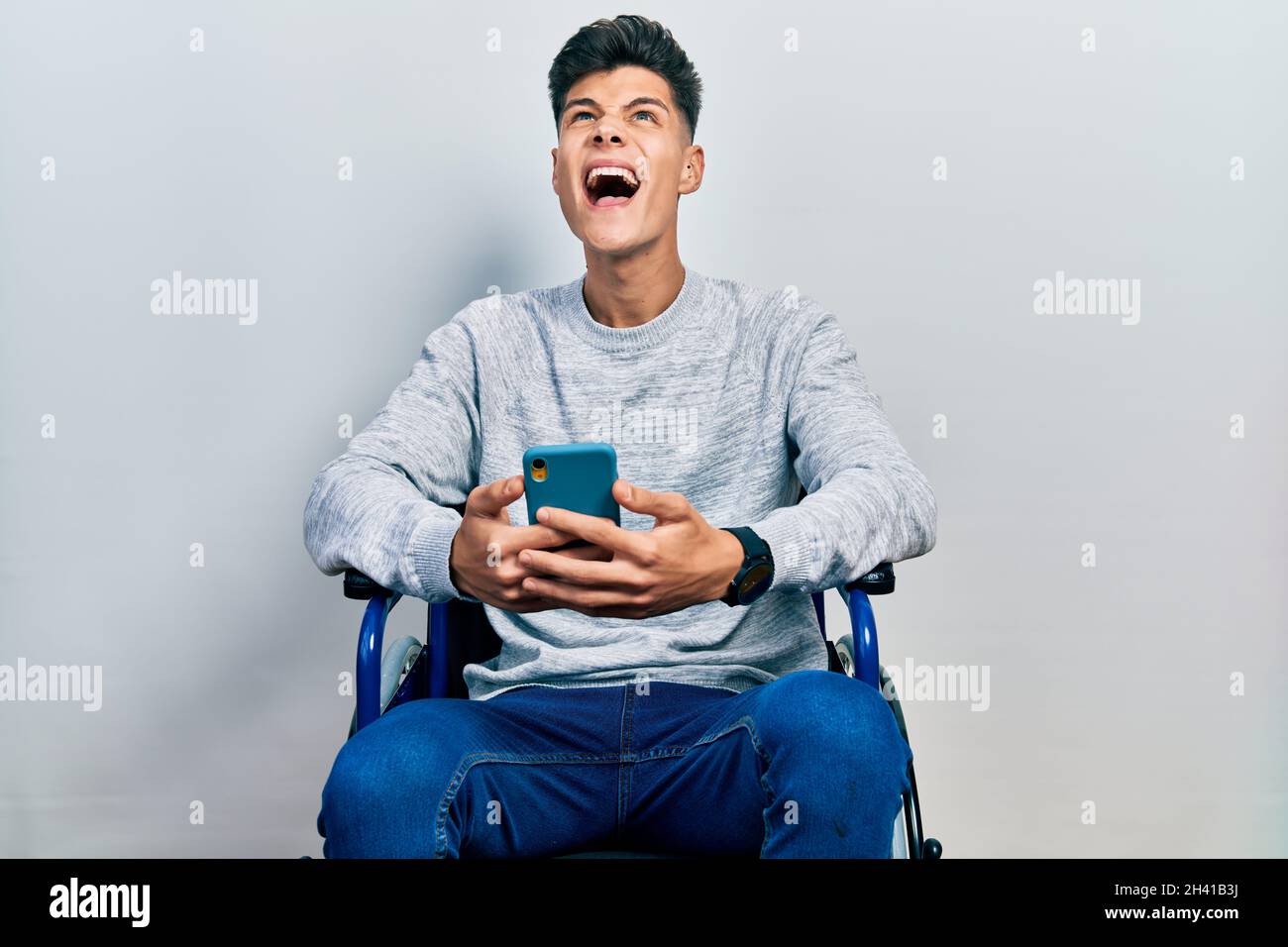 Young hispanic man sitting on wheelchair using smartphone angry and mad ...