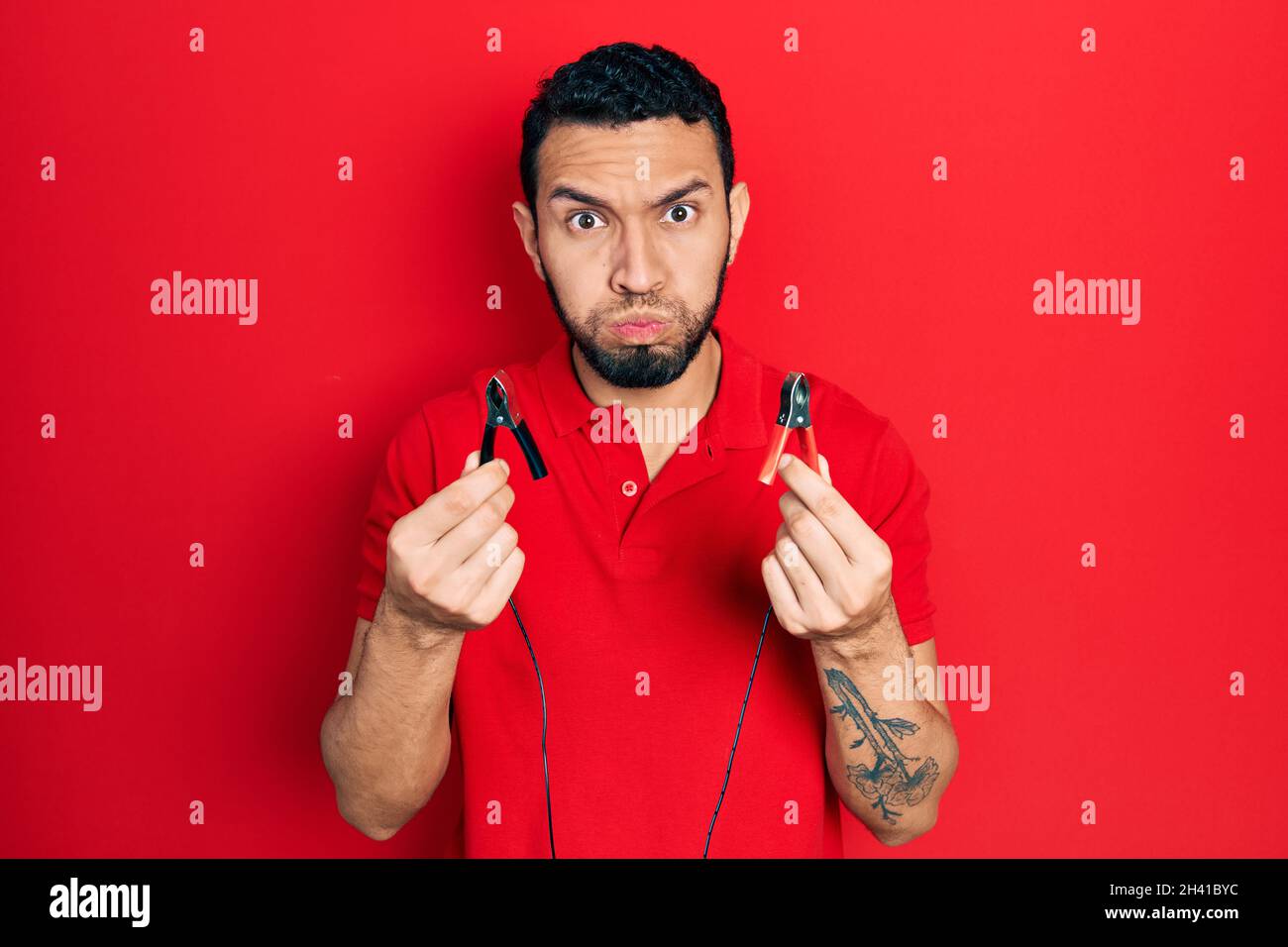Hispanic man with beard holding battery clamps puffing cheeks with ...