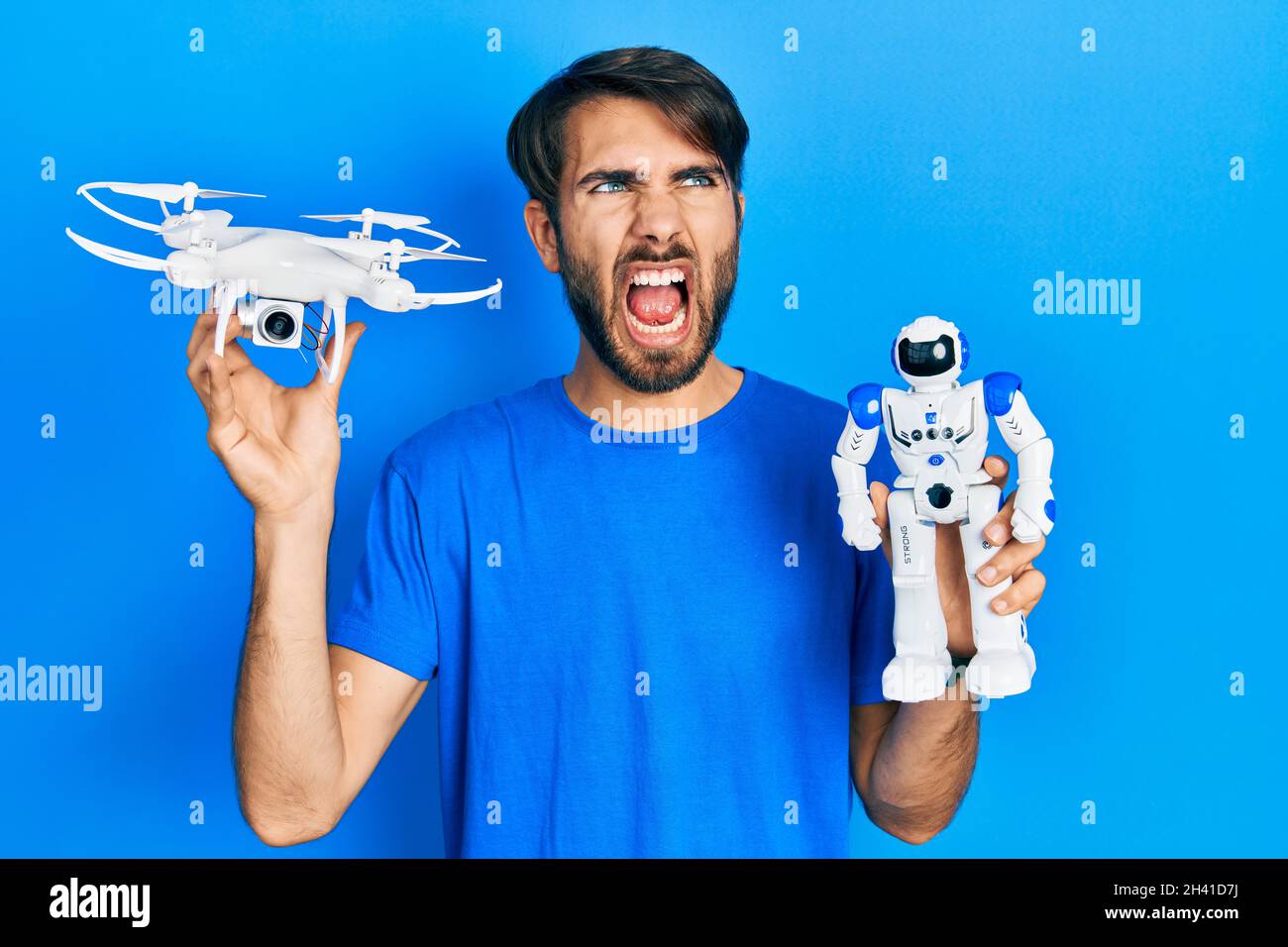 Young hispanic man holding drone and robot toy angry and mad screaming ...