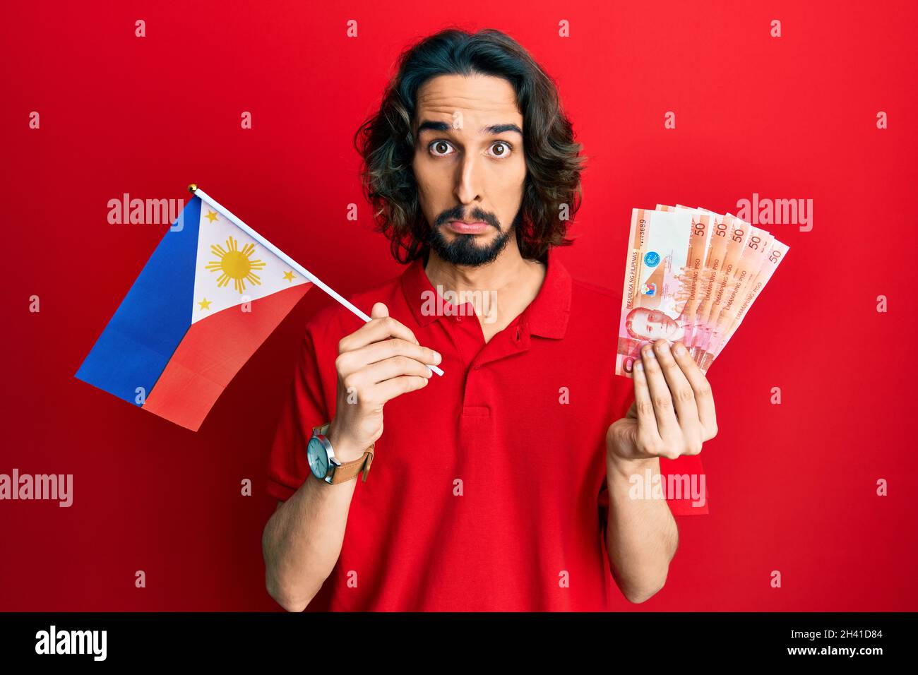Young hispanic man holding philippines flag and pesos banknotes ...