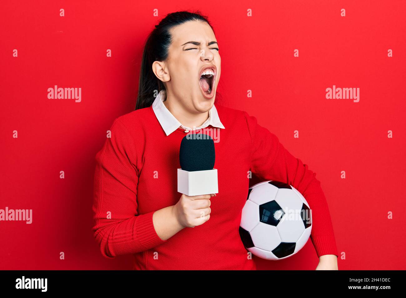 Young hispanic woman holding reporter microphone and soccer ball angry ...
