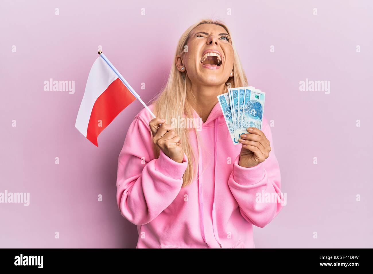 Young blonde woman holding poland flag and zloty banknotes angry and ...