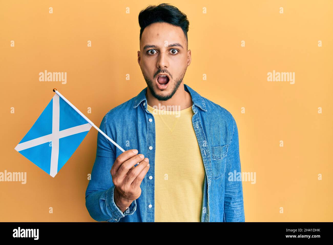 Young arab man holding scotland flag scared and amazed with open mouth ...