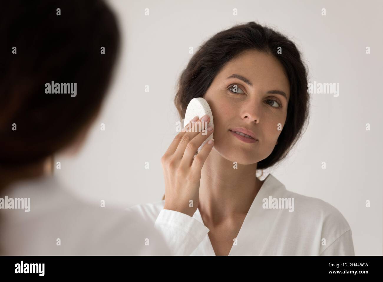 Beautiful woman reflected in mirror cleansing skin using facial sponge ...