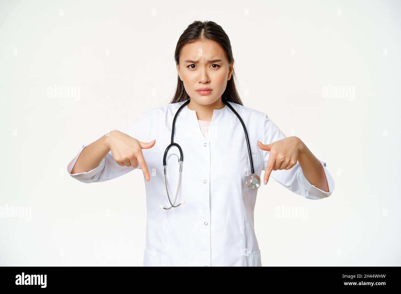Disappointed asian female doctor pointing fingers down, frowning and ...