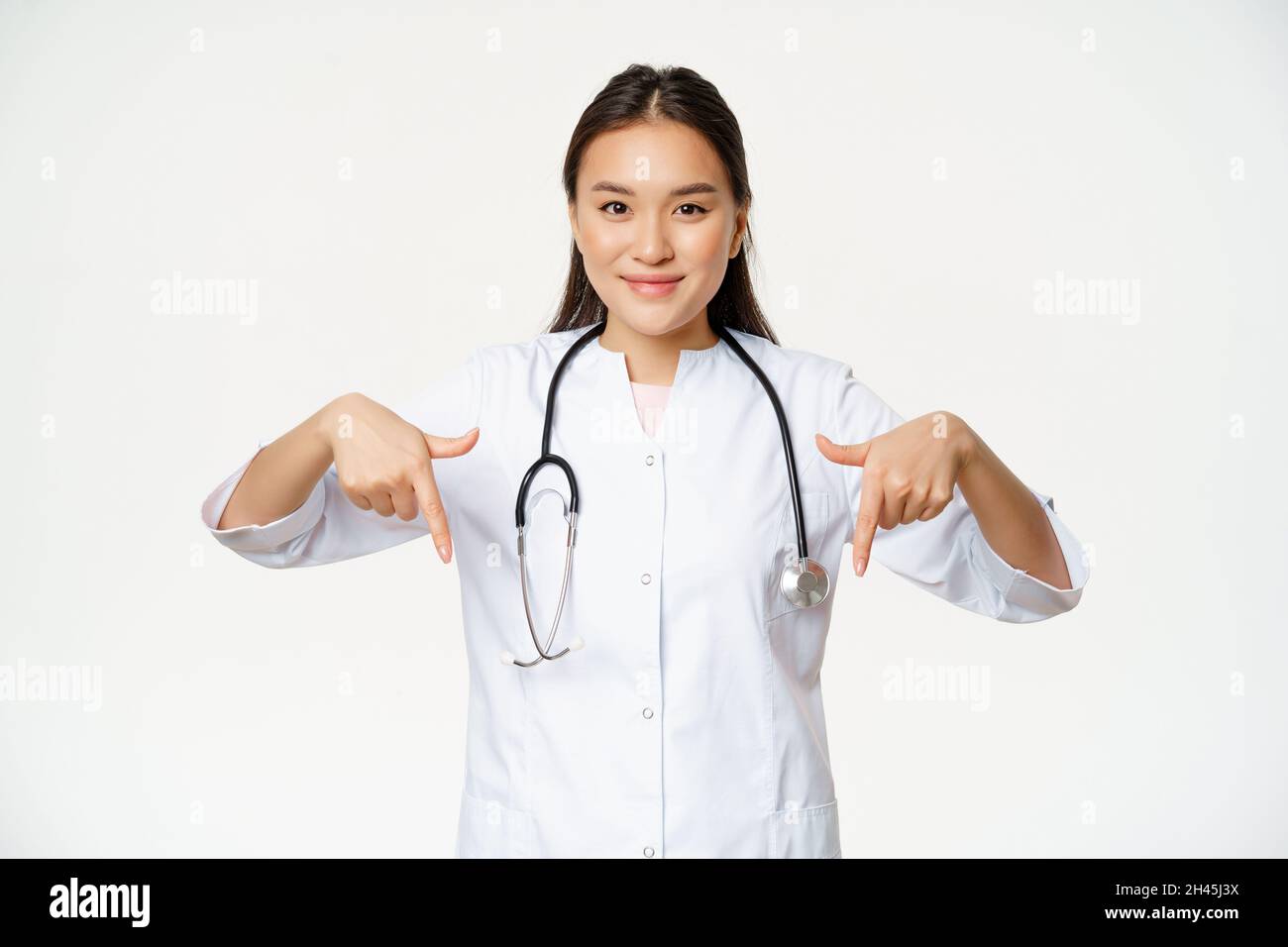 Young Asian Female Doctor Pointing Fingers Down And Smiling Wearing Stethoscope And Medical